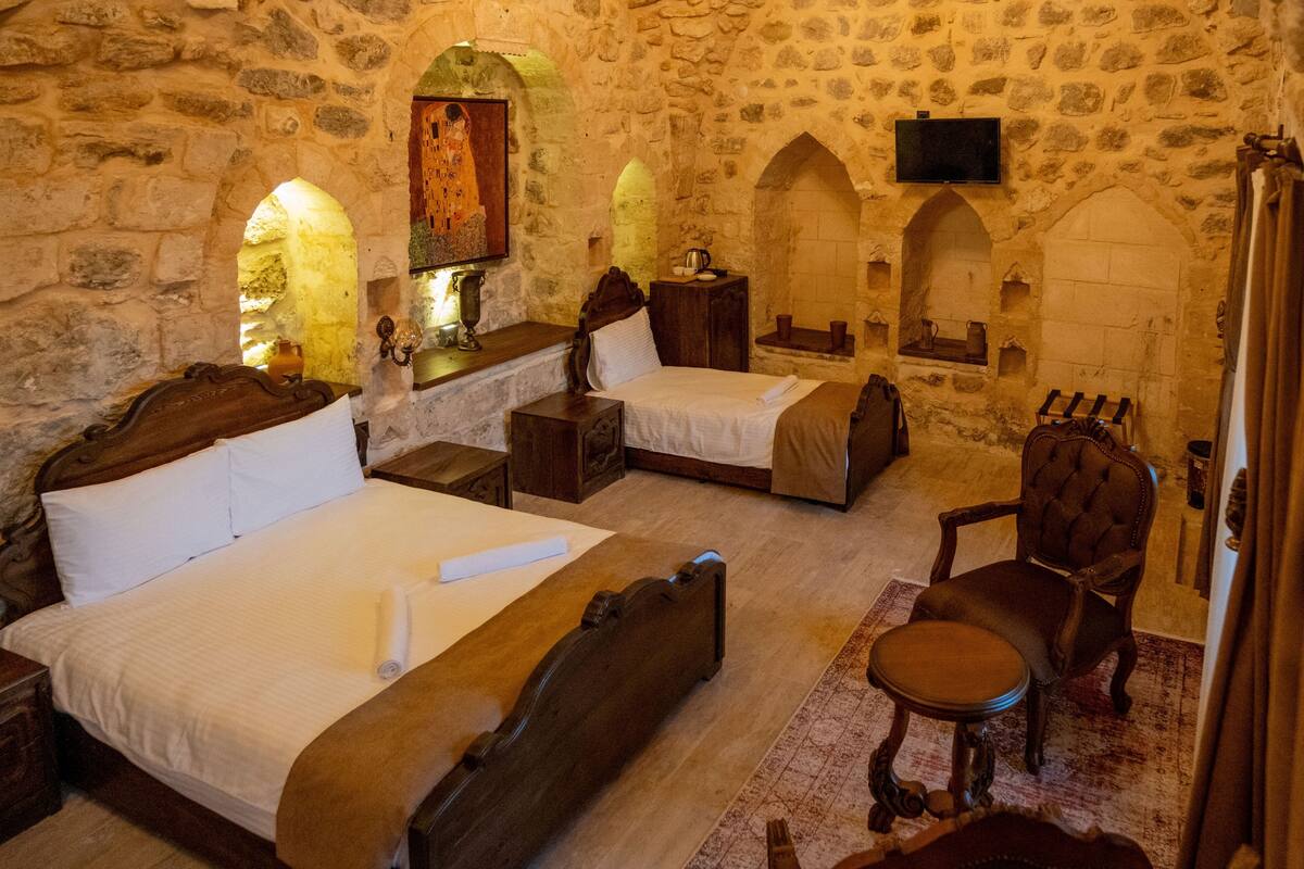 Quarto com paredes de pedra, duas camas, uma cadeira de madeira e uma televisão fixada na parede.