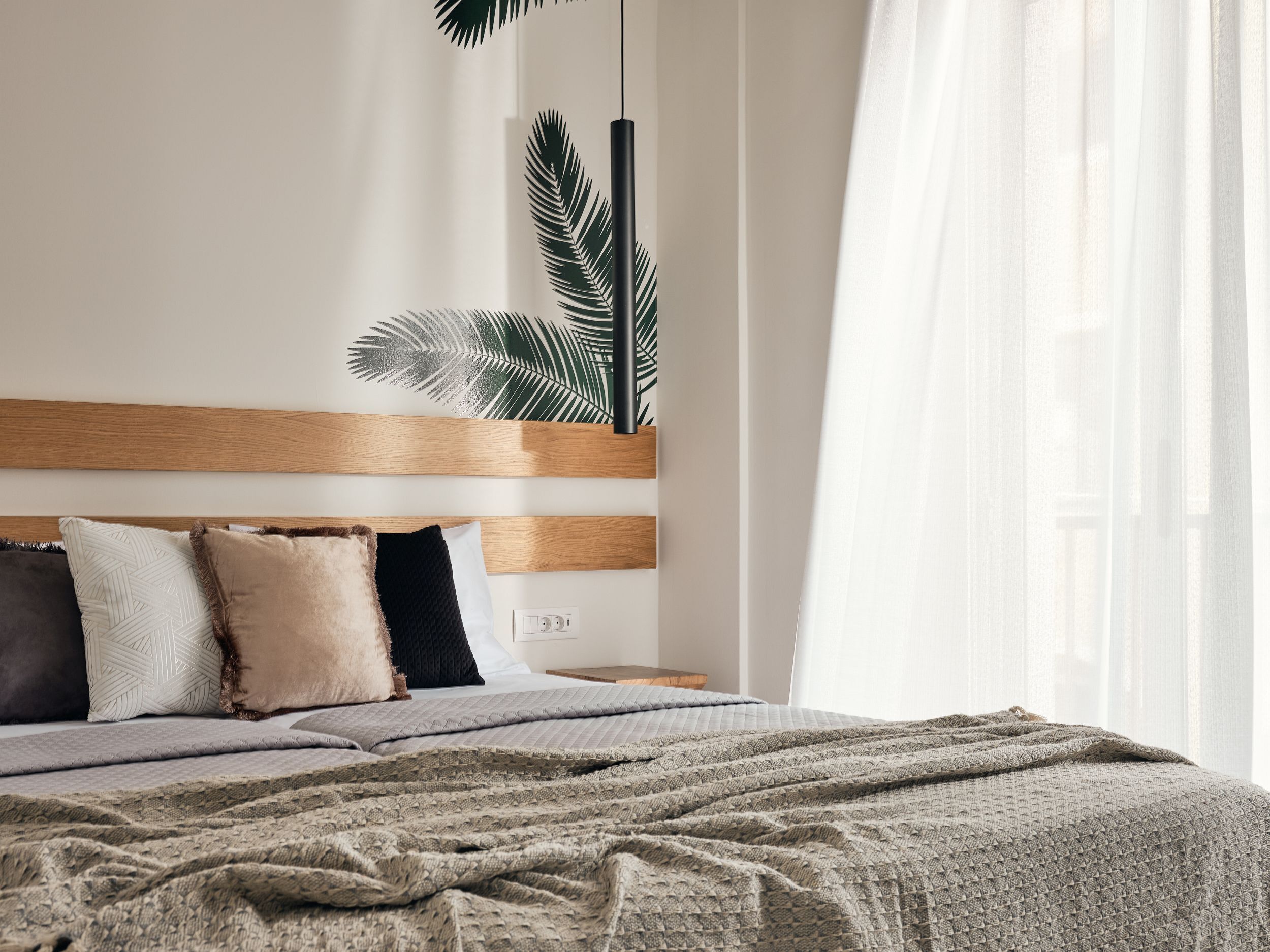 Cama bem arrumada com manta cinza e travesseiros, painel de madeira e planta fixada na parede.