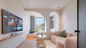 Quarto de hotel moderno com uma televisão de tela plana grande, um sofá, uma mesinha redonda e vista para a paisagem urbana através de uma janela em arco.
