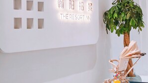 Interior de hotel com parede branca exibindo um logotipo e nome, um vaso de planta e uma área de recreação com brinquedos e uma mesa pequena.