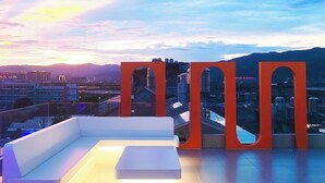 Terraço no topo do prédio com um sofá branco e detalhes arquitetônicos vermelhos, com vista para o horizonte urbano ao pôr do sol.