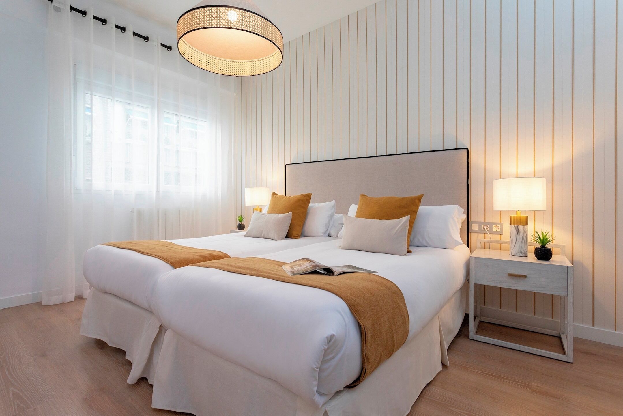 Uma cama de casal com roupa de cama branca e castanha, uma mesa de cabeceira com abajur e uma janela com cortinas leves.