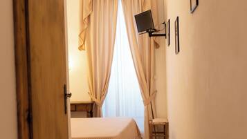 Um corredor estreito com uma porta de madeira, uma cama com colcha estampada, uma cadeira e uma TV fixada na parede.