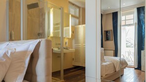 Quarto moderno com chuveiro em cabine de vidro, mesa de cabeceira, cama e vista para o exterior através de uma janela.