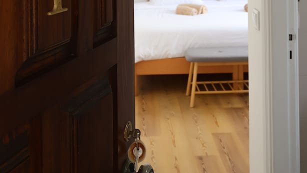Quarto de hotel com porta de madeira, uma cama com roupa de cama branca, um banco de madeira e uma pintura colorida na parede.