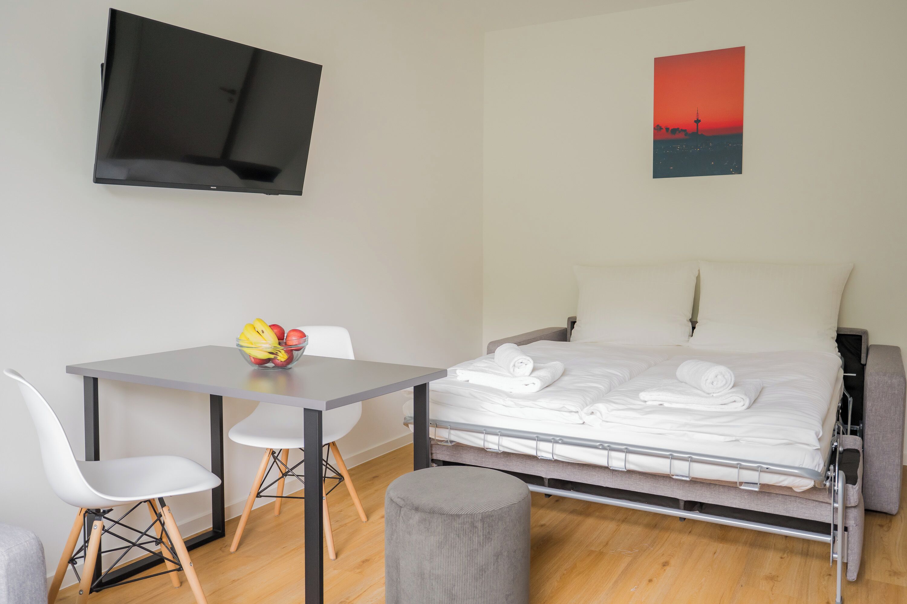 Quarto de hotel com cama, escrivaninha, duas cadeiras, televisão e um quadro na parede.