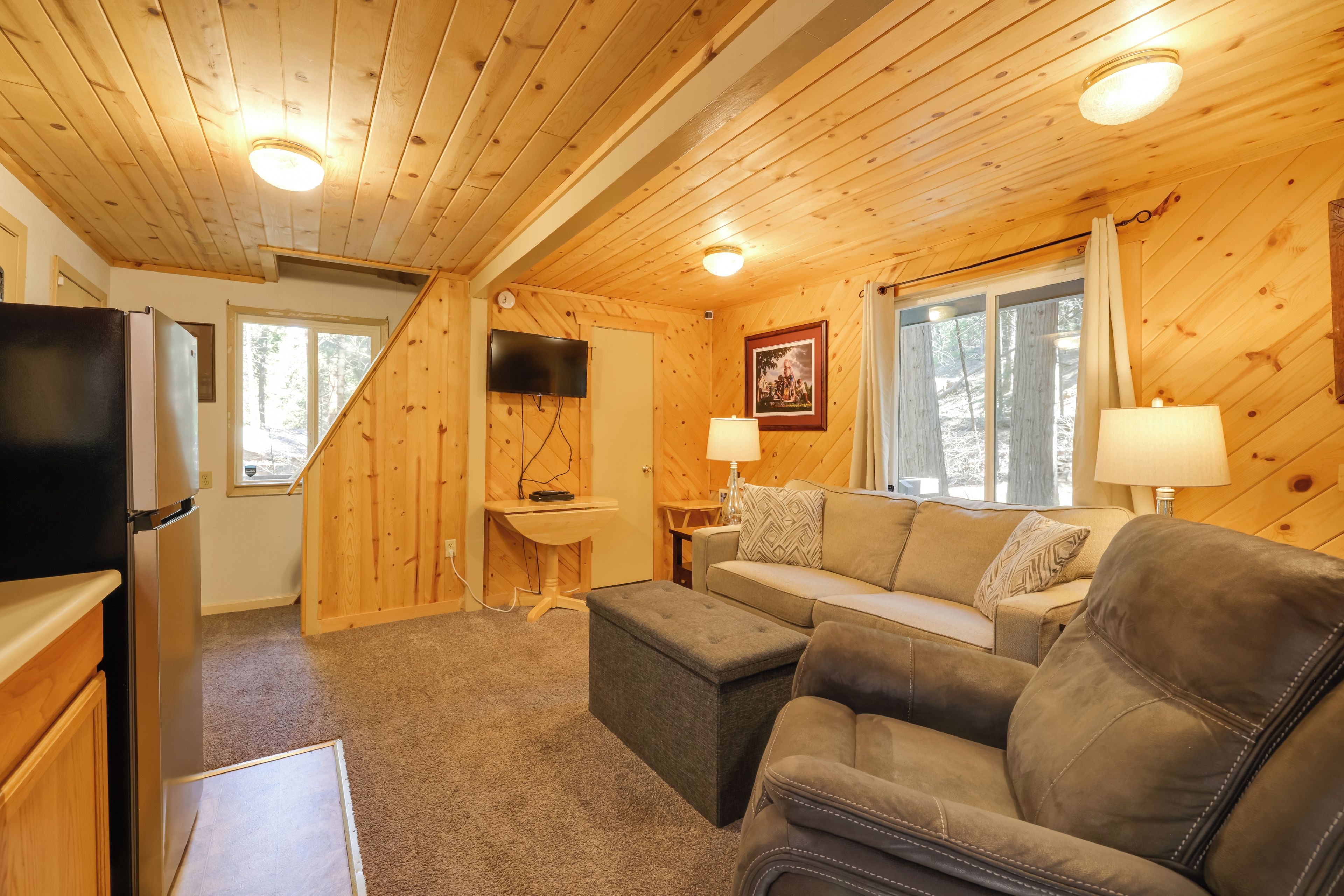 Interior acolhedor de uma cabana, com paredes de madeira, um sofá, uma televisão e uma janela com cortinas.