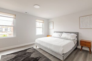Quarto moderno com uma cama grande, duas mesas de cabeceira e vista da cidade por grandes janelas.