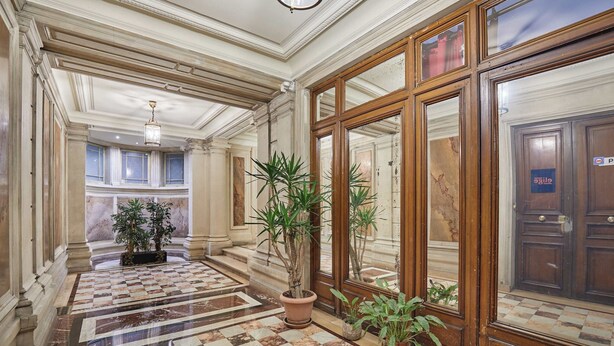 Um corredor com piso de mármore quadriculado, portas de madeira grandes e plantas em vasos.