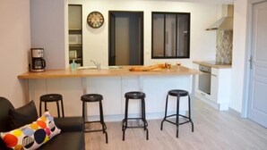 Cozinha compacta com ilha central, micro-ondas e máquina de café. Três bancos na ilha e um relógio fixado na parede.