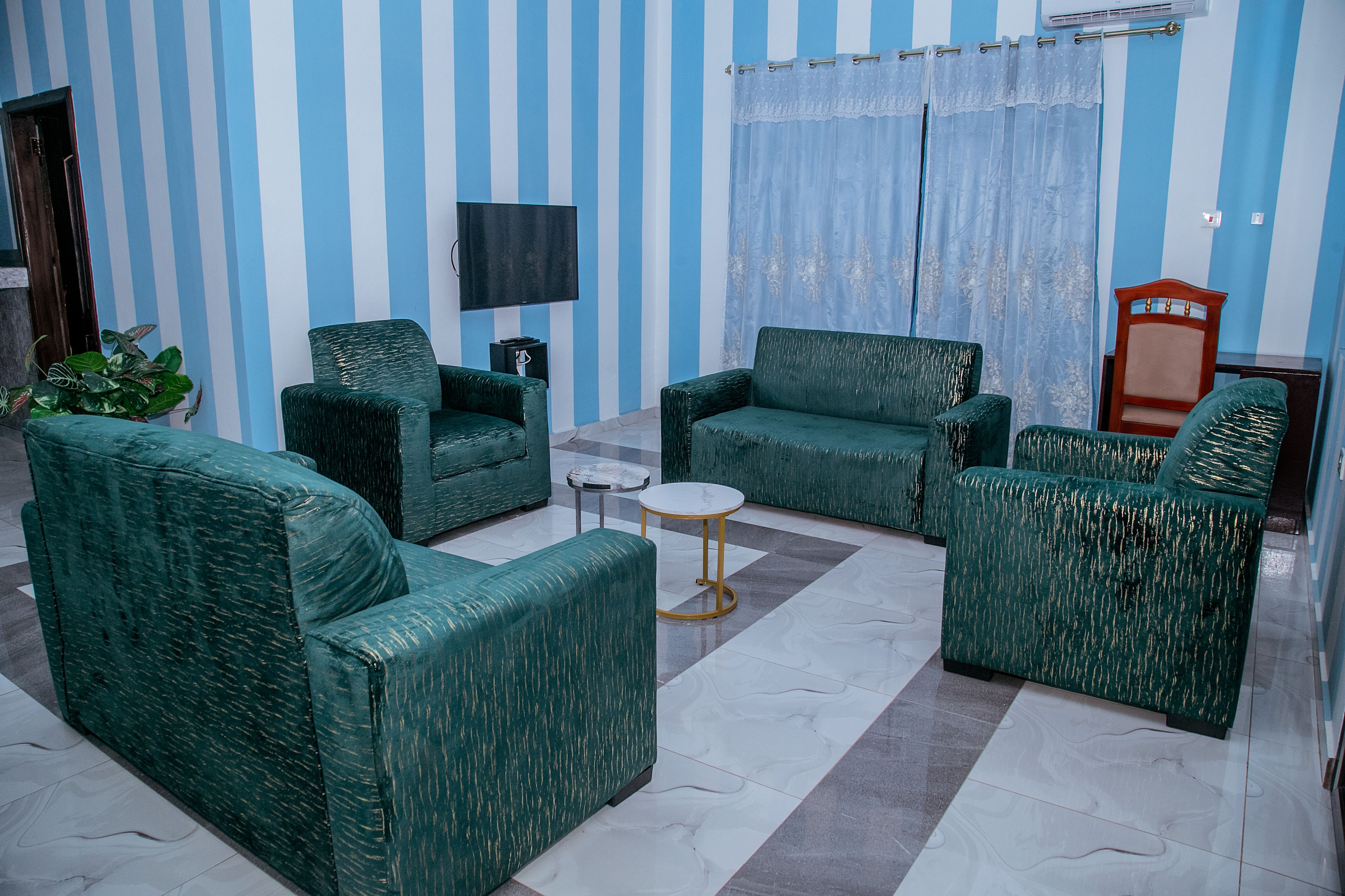 Uma sala de estar com paredes listradas em azul e branco, uma televisão de tela plana na parede, um sofá, duas poltronas, uma mesinha de centro redonda e um vaso com planta.