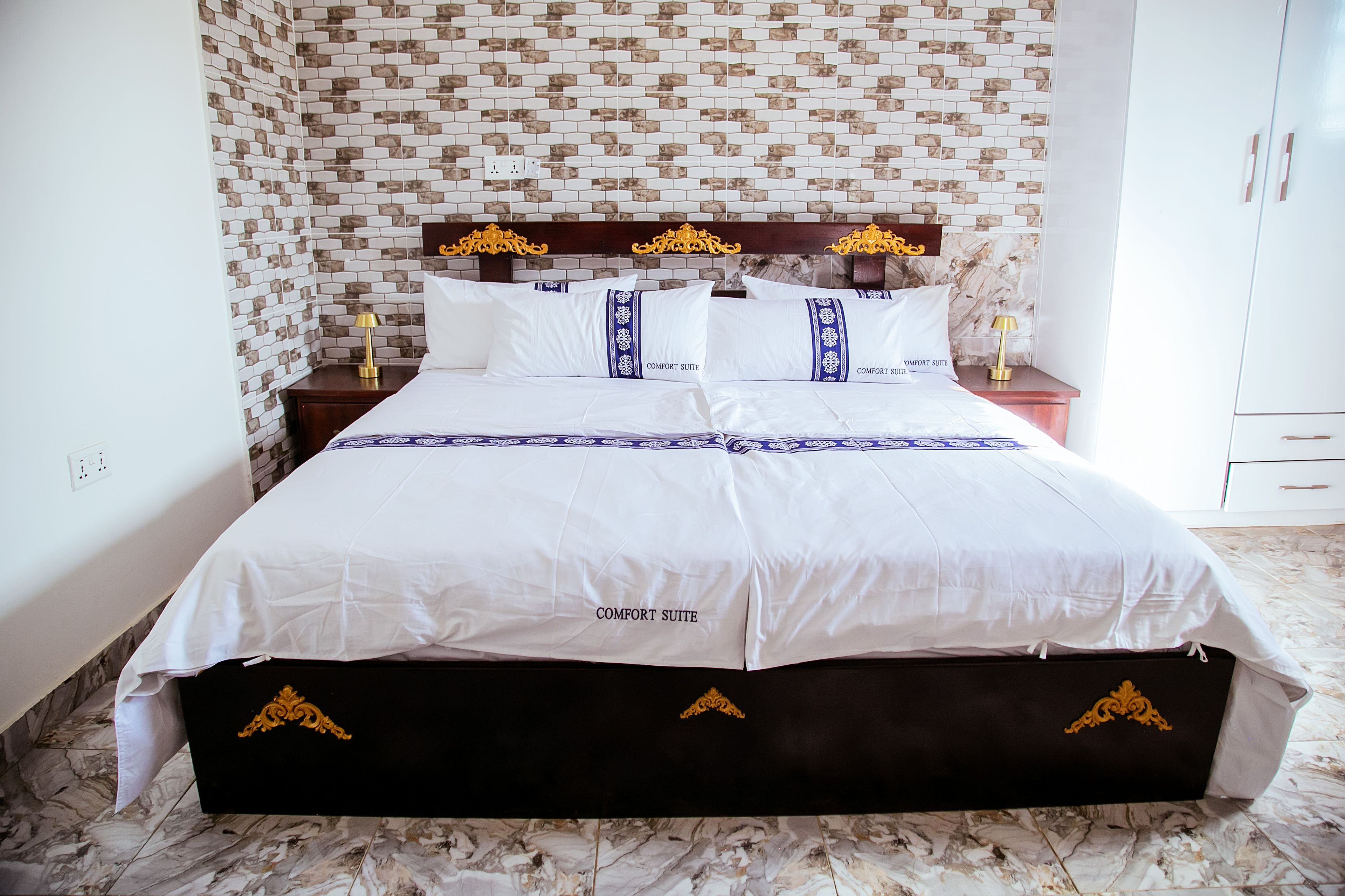 Uma cama com colcha branca e confortável, cabeceira de madeira e piso de mármore.