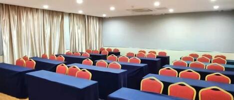 Sala de conferências com mesas e cadeiras dispostas em fileiras, um projetor e uma tela grande.