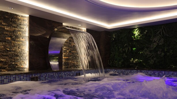 Uma banheira de hidromassagem com cascata, cercada por um muro de pedra e um piso revestido de azulejos.