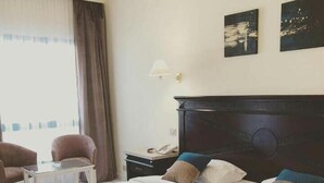Quarto de hotel com uma cama grande, mesa de cabeceira, uma cadeira e janela com cortinas.