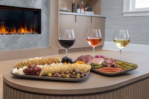 Cozinha bem iluminada com lareira moderna, variedade de queijos, bolachas e charcutaria em uma bandeja, e dois copos de vinho, um com tinto e outro com branco.