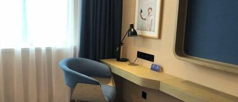 Quarto de hotel com escrivaninha, cadeira, janela com cortinas, um quadro emoldurado e uma janela ampla.