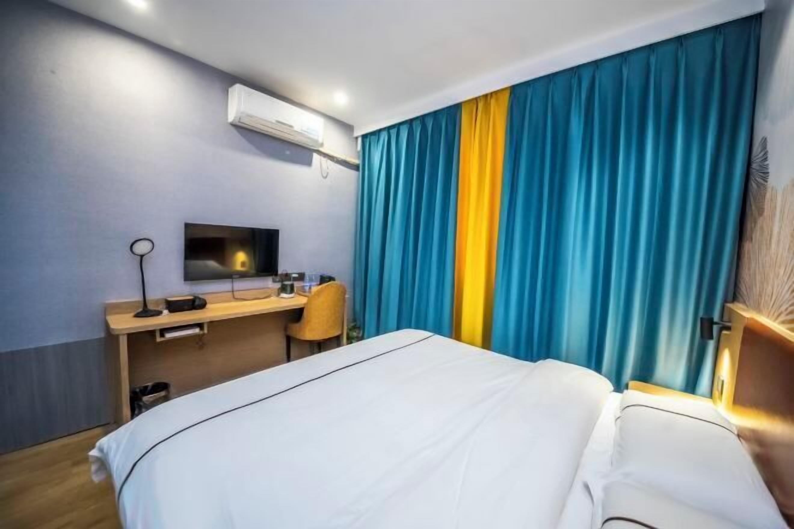 Quarto de hotel com cama, escrivaninha com cadeira, televisão e janela com cortinas.