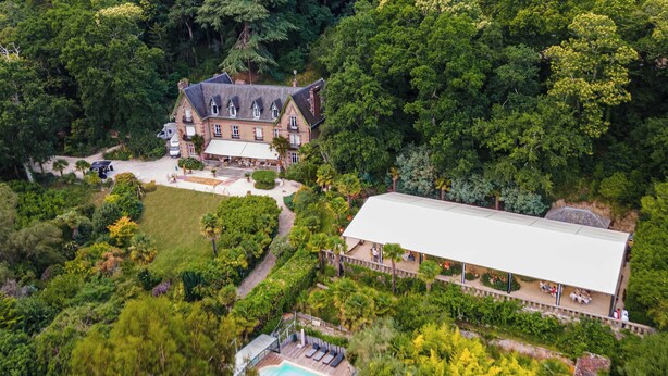 Vista aérea de um grande edifício histórico cercado por vegetação exuberante, com piscina e área de estar ao ar livre.