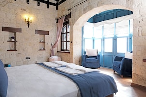 Quarto com uma cama grande, duas cadeiras, parede de pedra, janela com cortinas e um candelabro.