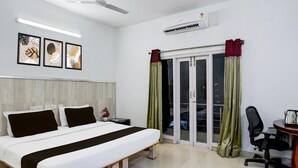 Quarto de hotel com cama, escrivaninha com cadeira, janela com cortinas e arte nas paredes.