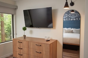 Quarto com TV de tela plana na parede, cômoda de madeira com gavetas, um pequeno vaso de planta e cama com cabeceira estampada.