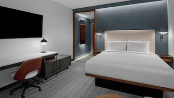 Quarto de hotel moderno com uma cama grande, uma escrivaninha com cadeira, uma luminária de mesa e uma pequena mesa redonda.