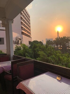 Uma área externa com mesas e cadeiras, com vista para um horizonte urbano com um edifício imponente e palmeiras.