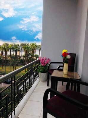 Uma varanda com mesa e cadeiras, um vaso com flores e vista para palmeiras e um corpo d'água.