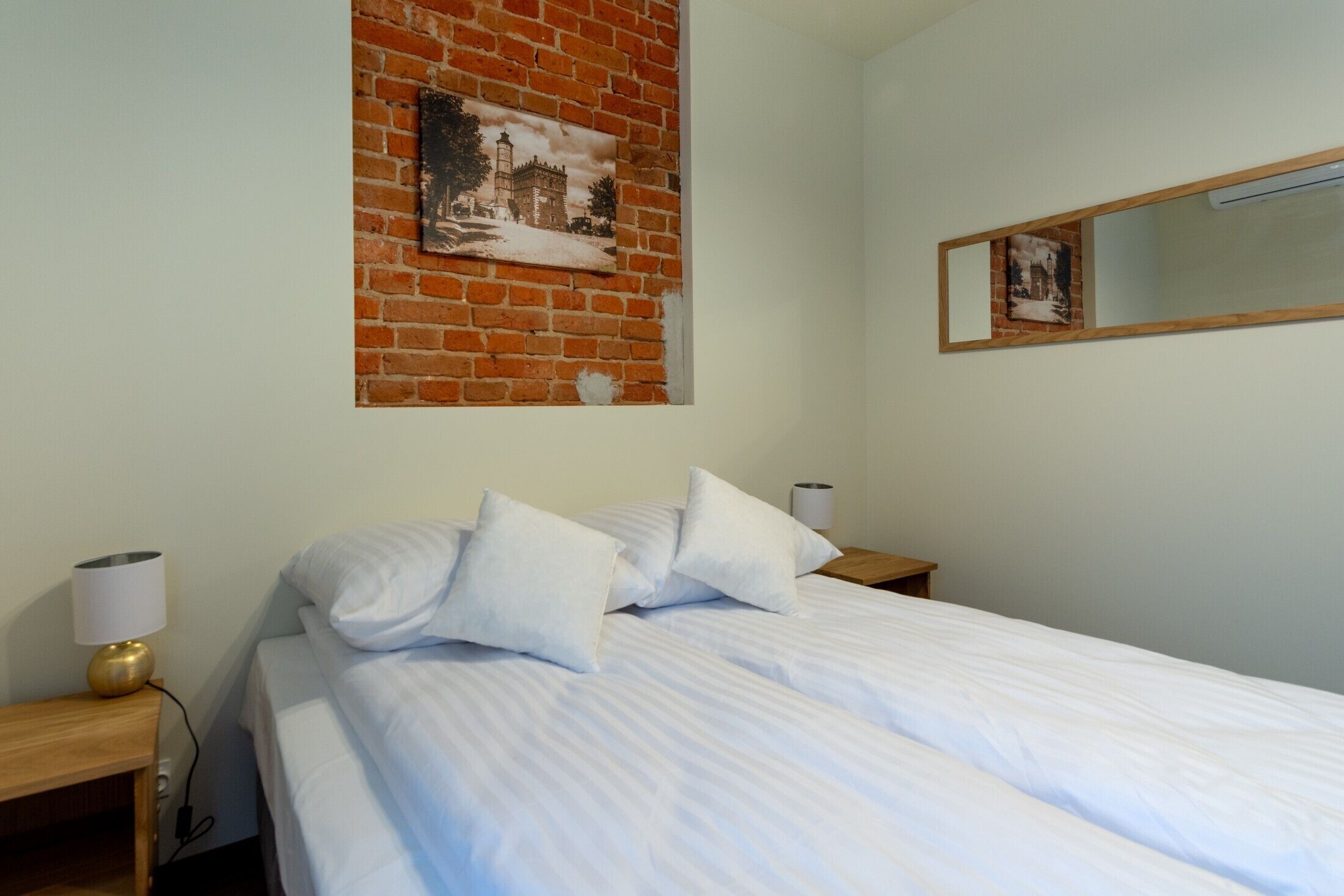 Quarto com uma cama de casal, roupa de cama branca, parede de tijolos com um quadro, mesa de cabeceira de madeira com uma luminária e um espelho na parede.