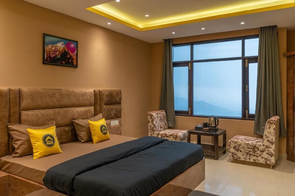 Quarto de hotel com uma cama grande, duas poltronas, uma mesa pequena e uma janela ampla com vista para as montanhas.