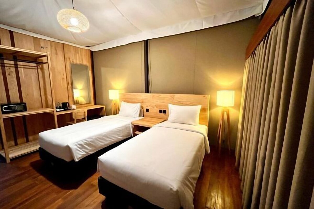 Quarto de hotel com duas camas de solteiro, um guarda-roupa de madeira, uma escrivaninha com cadeira e abajures nas laterais.