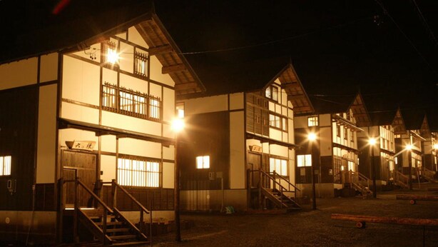 Uma fileira de edifícios em estilo japonês tradicional à noite, com janelas iluminadas e escadas de madeira.