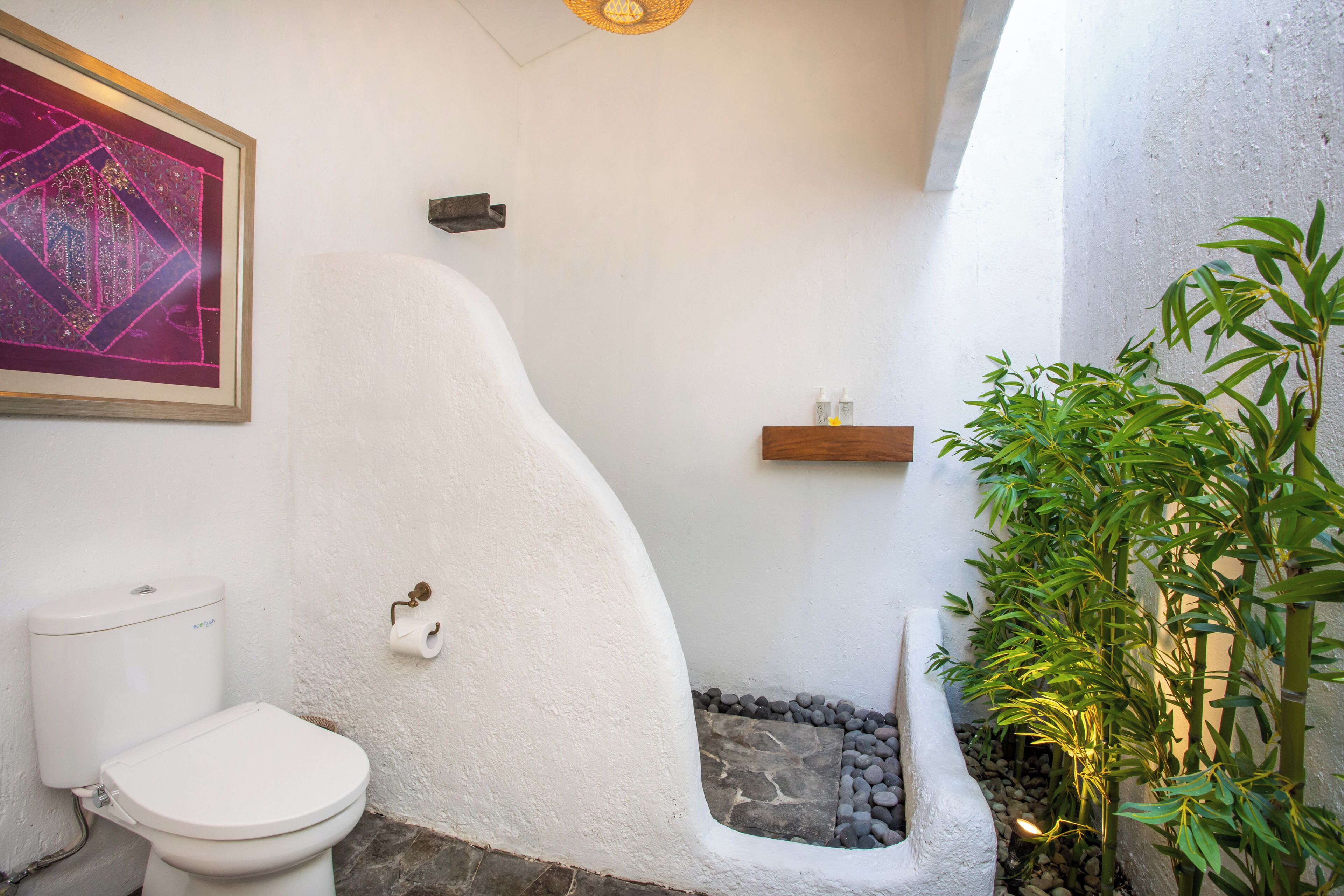 Casa de banho com sanita branca, uma obra de arte emoldurada, lavatório de pedra e planta de bambu.
