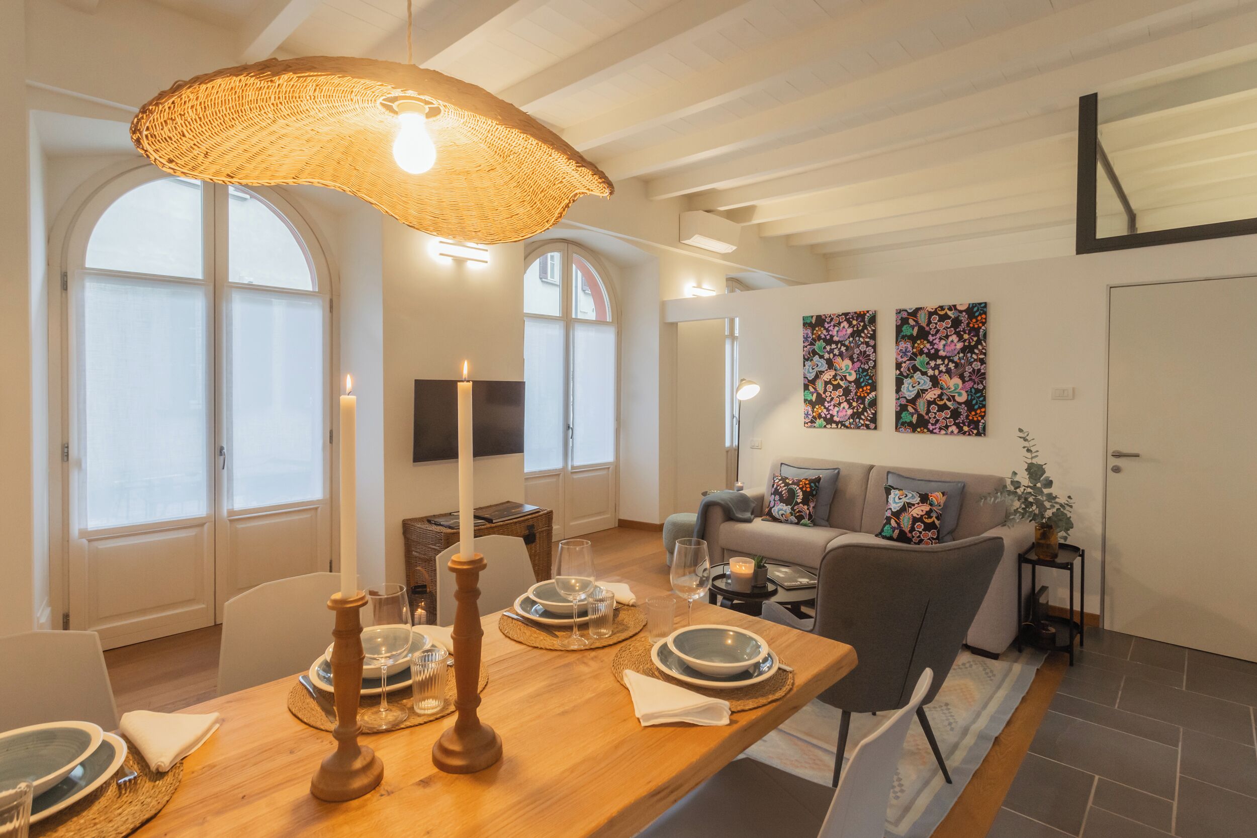 Uma área de refeições com uma mesa de madeira posta para quatro pessoas, uma luminária pendente trançada e uma vela.