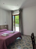 Quarto de hotel com cama, uma cadeira, janela com cortinas e vista para o verde lá fora.