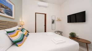 Quarto de hotel com uma cama, uma televisão, uma mesinha de cabeceira com uma planta e uma porta.