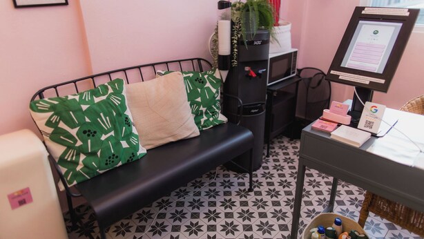 Quarto com um banco de metal preto, almofadas com padrão verde, uma pequena escrivaninha com um computador e chão de azulejos.
