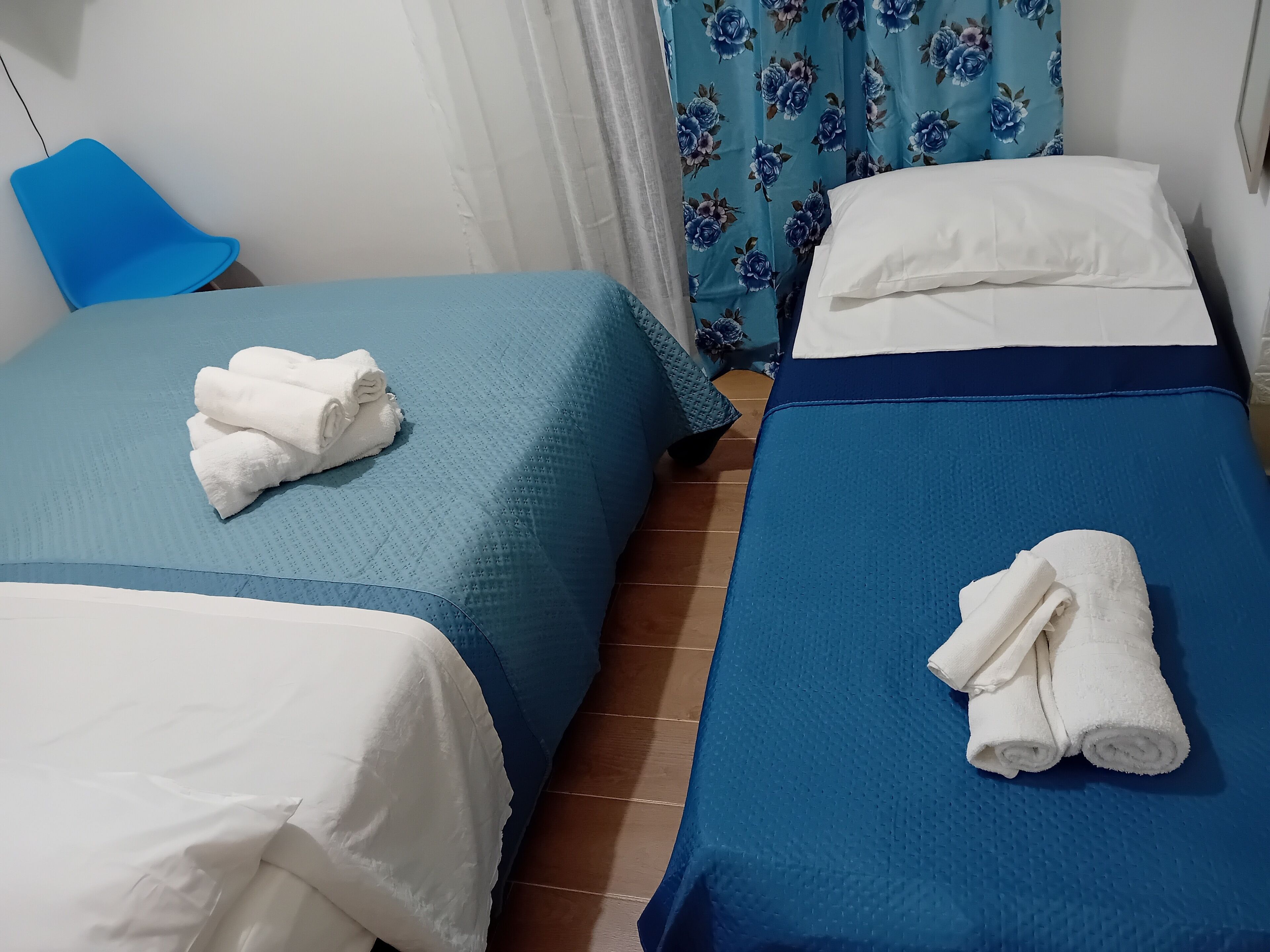 Duas camas de solteiro com roupa de cama azul e branca, uma cadeira azul e uma cortina florida.