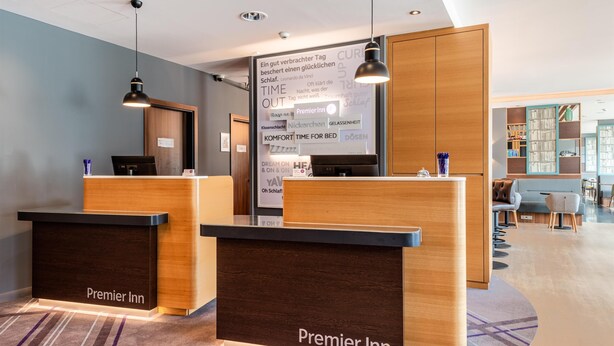 Uma área de recepção de hotel com dois balcões identificados como 'Premier Inn', uma parede grande com diversos serviços listados e um espaço de espera ao fundo.