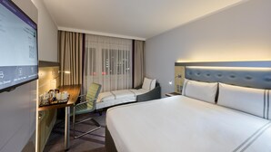 Quarto de hotel com uma cama grande, uma escrivaninha com cadeira, televisão e janela com cortinas.
