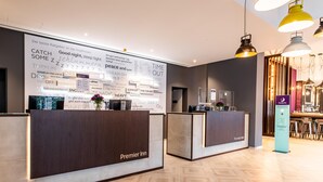 Uma área de recepção de hotel com dois balcões de recepção identificados como 'Premier Inn', uma parede com várias palavras e frases, e um espaço bar com assentos.