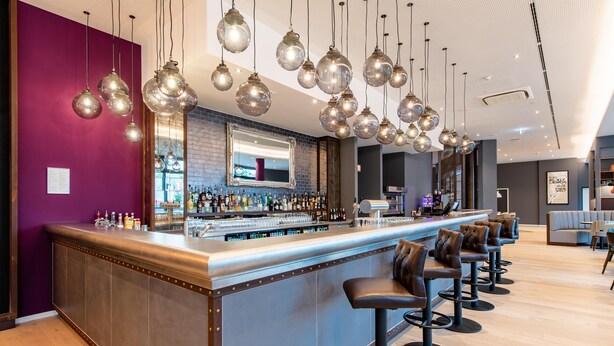 Uma área de bar com um balcão comprido, várias luminárias pendentes e um espaço de estar com banquetas.
