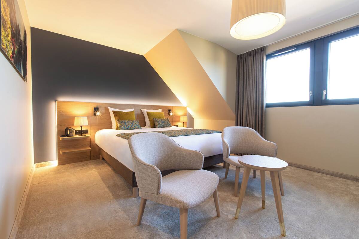 Quarto de hotel moderno com uma cama grande, duas poltronas, uma mesinha, uma mesa de cabeceira com abajur e uma janela ampla com cortinas.