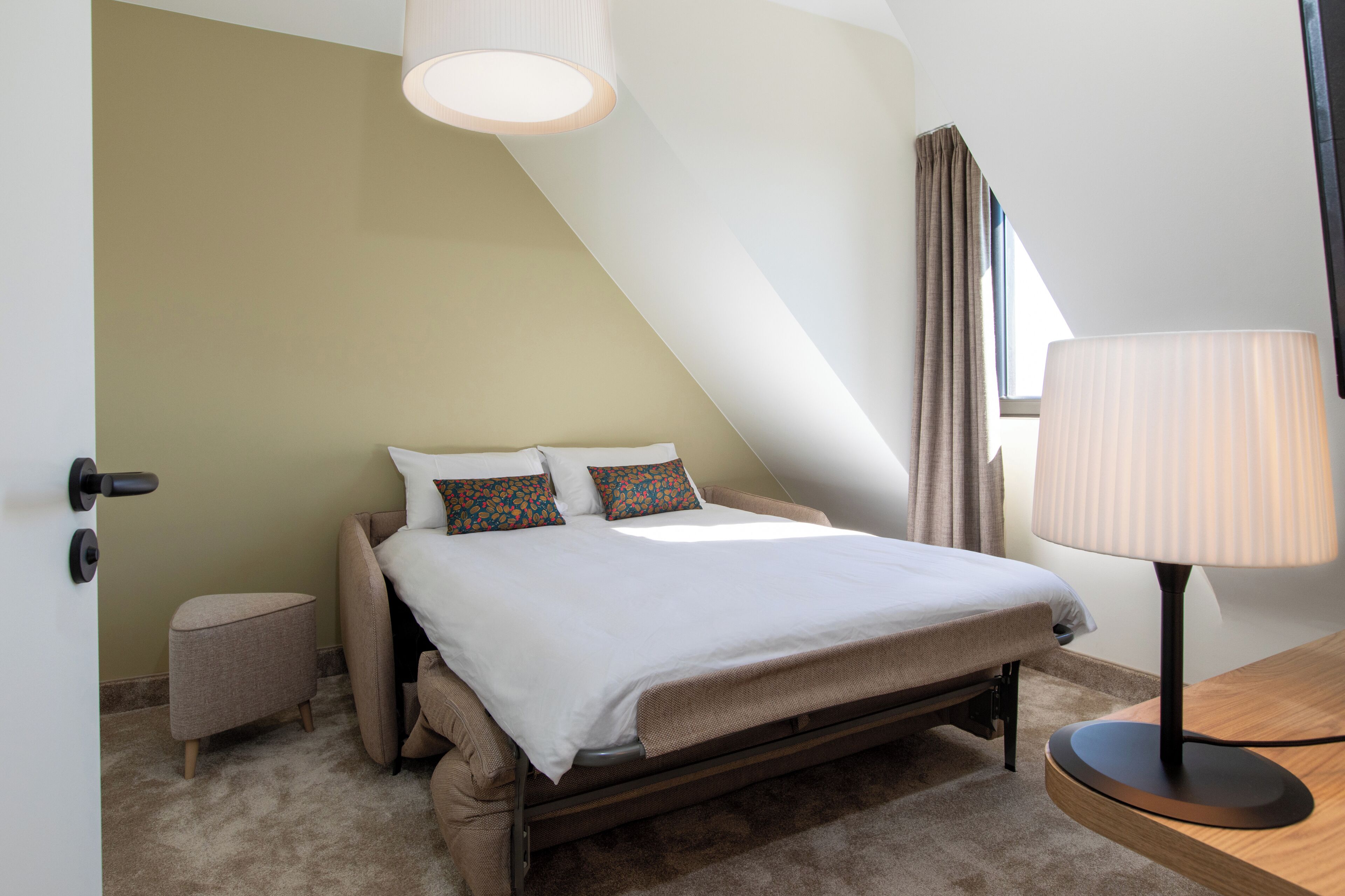 Quarto com cama, mesa de cabeceira, abajur e um banquinho.