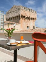 Uma mesa com itens de café da manhã, incluindo uma xícara, um copo de suco de laranja, um doce e uma planta. Ao fundo, um edifício de pedra com uma torre.