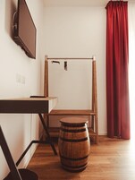 Quarto com escrivaninha de madeira, cadeira de madeira, banquinho de madeira, estrutura de cama de madeira, cortina vermelha e televisão montada na parede.