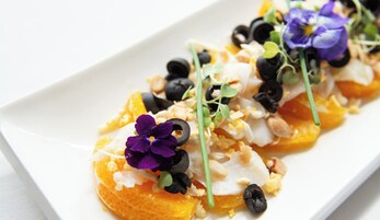 Um prato retangular branco com uma apresentação de laranja em gomos, flores roxas, azeitonas pretas e brotos verdes.