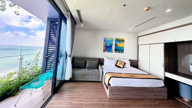 Quarto moderno com uma grande janela com vista para o mar, uma cama com cabeceira de madeira, um sofá e uma pequena varanda com uma rede.