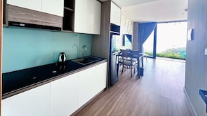 Cozinha moderna com armários brancos, bancada preta e fogão embutido. Há uma área de refeições com mesa e cadeiras, e uma varanda com vista.